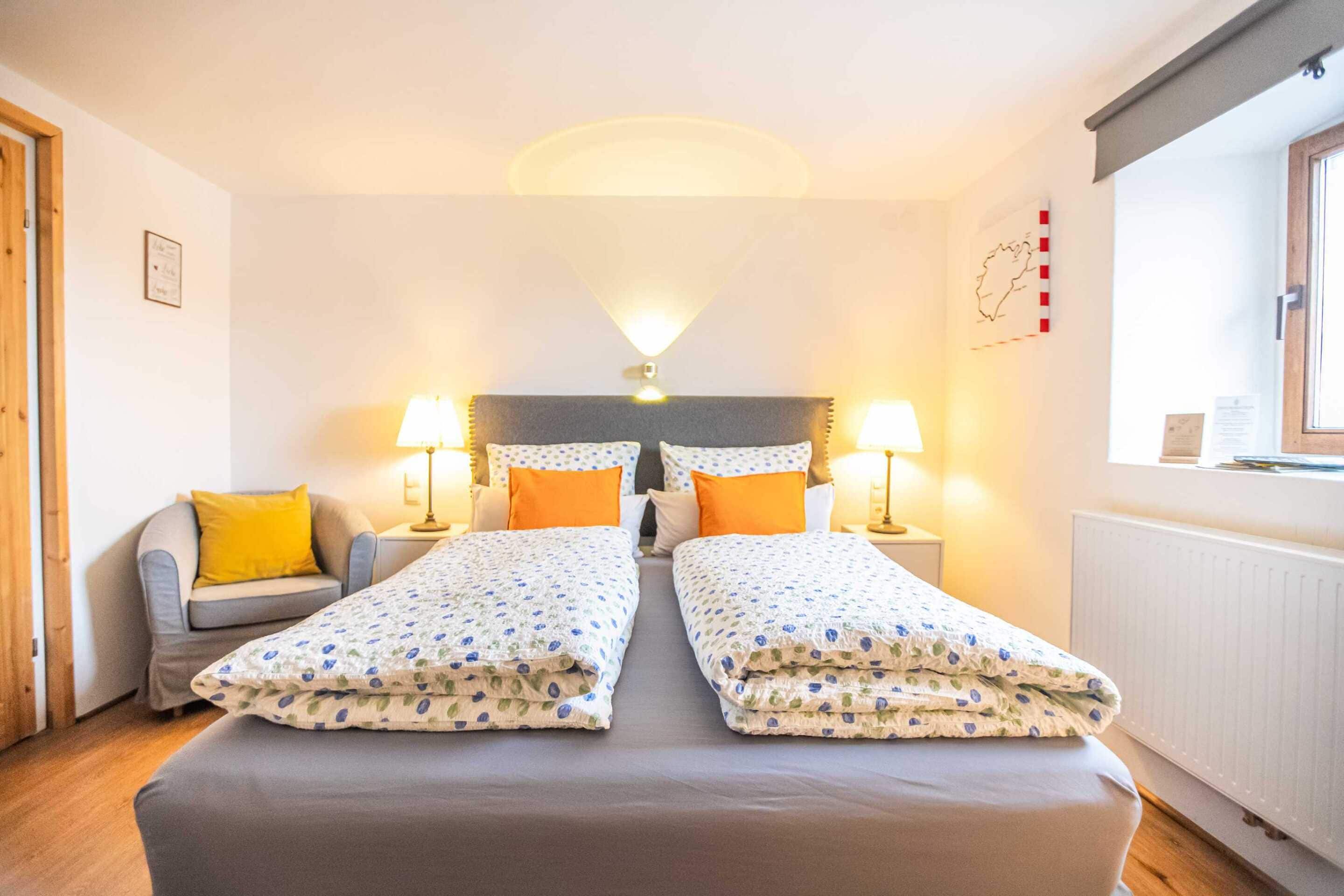Uma cama de casal com colcha florida, duas luminárias de cabeceira, um sofá cinza com almofada amarela e uma janela com um mapa na parede.