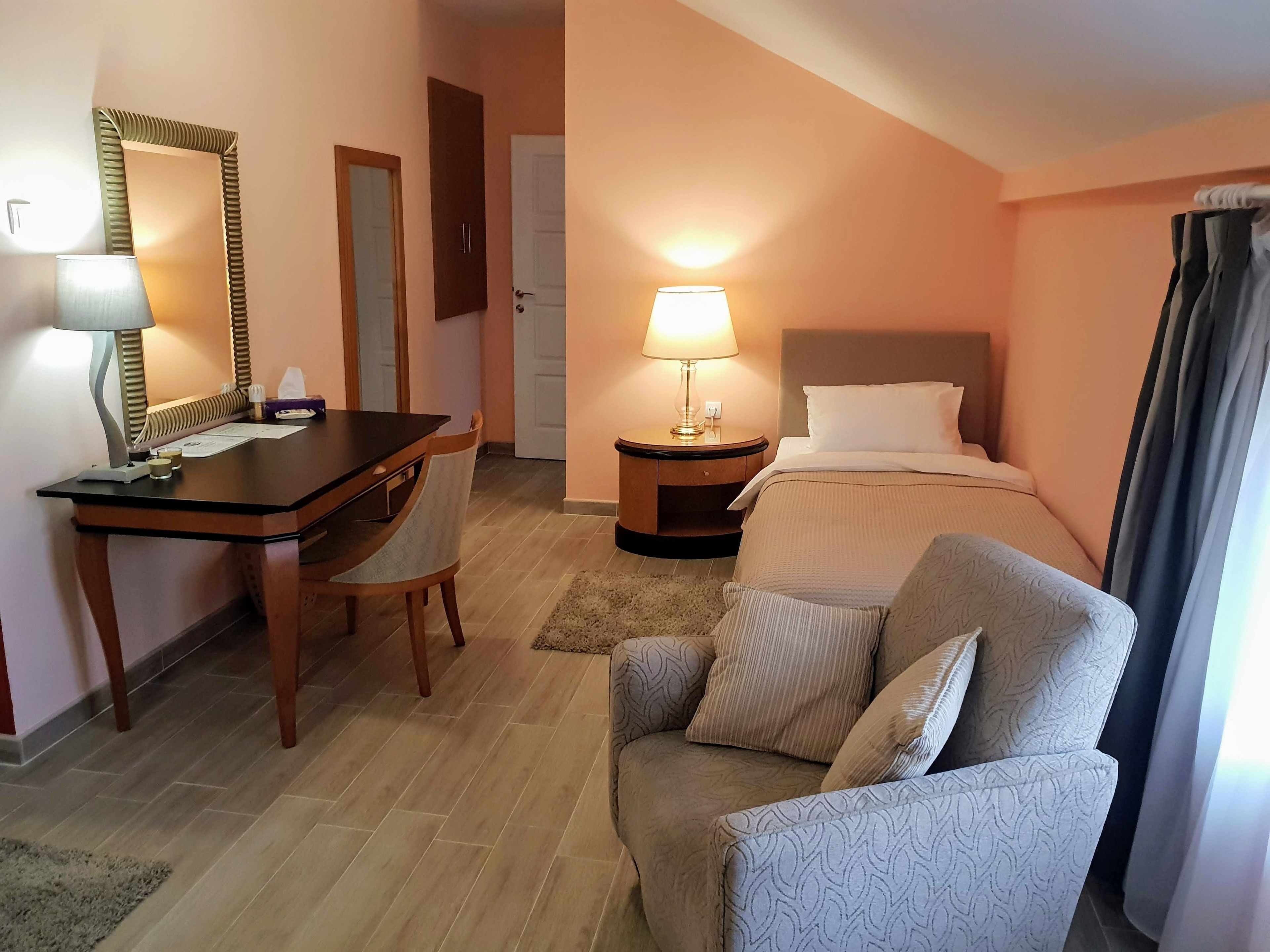 Quarto de hotel com cama, escrivaninha, cadeira, mesa de cabeceira com abajur e um espelho grande.
