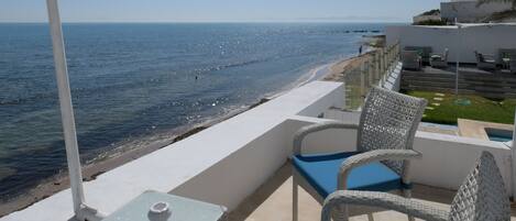 Varanda com cadeiras de vime e uma mesa, com vista para a praia e o mar.