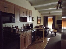 Cozinha com armários de madeira, micro-ondas e cafeteira. Há uma área de jantar com mesa e cadeiras, e uma sala de estar com sofá e televisão.