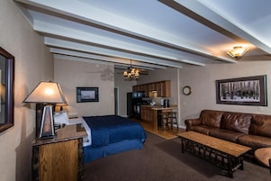 Quarto de hotel com cama, escrivaninha, mesa de centro, sofá, cozinha compacta e ventilador de teto.