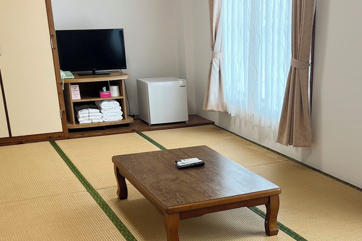 Quarto com tatames, uma mesa de madeira, televisão, um pequeno frigorífico e uma prateleira com toalhas.