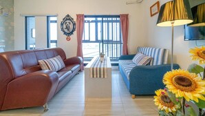 Sala de estar com um sofá vermelho, um sofá azul, uma mesa de centro e um abajur. Há girassóis no chão e uma janela com cortinas.