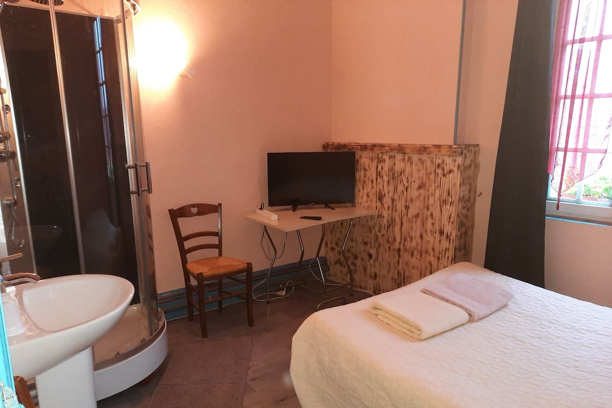 Um quarto pequeno com uma cama, uma escrivaninha com computador, uma cadeira, pia e chuveiro.