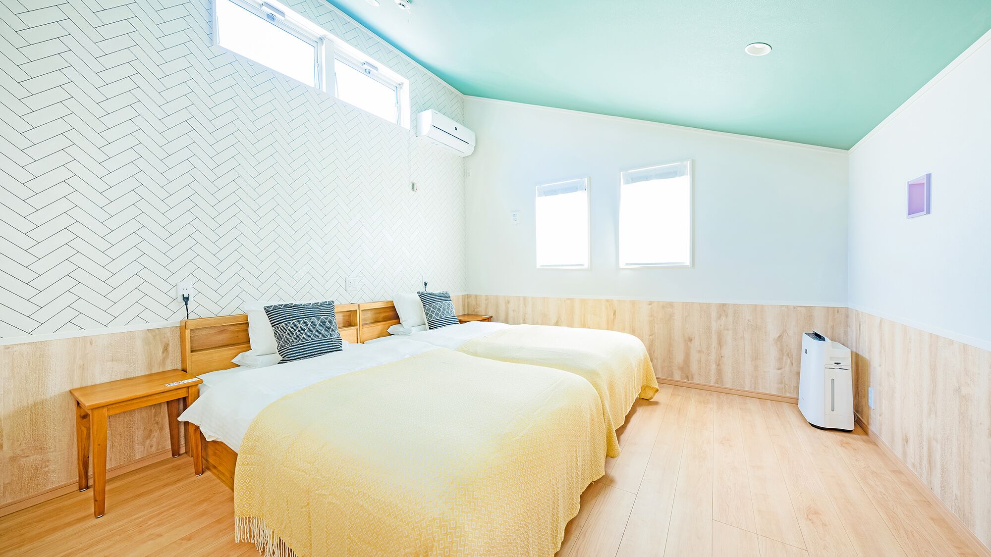 Quarto com duas camas, um banco de madeira, um ar condicionado branco e parede com padrão herringbone.