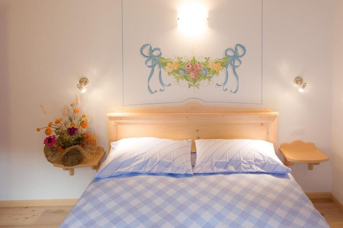 Quarto com cabeceira de madeira, colcha xadrez, luminária na parede e arranjo floral decorativo em uma prateleira.