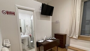 Quarto de hotel com piso de madeira, uma cama, uma mesa com cadeira, televisão na parede e uma janela com cortinas.