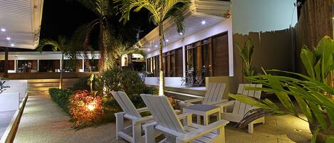 Uma área externa bem iluminada com cadeiras Adirondack brancas e uma mesa, rodeada por palmeiras e plantas.