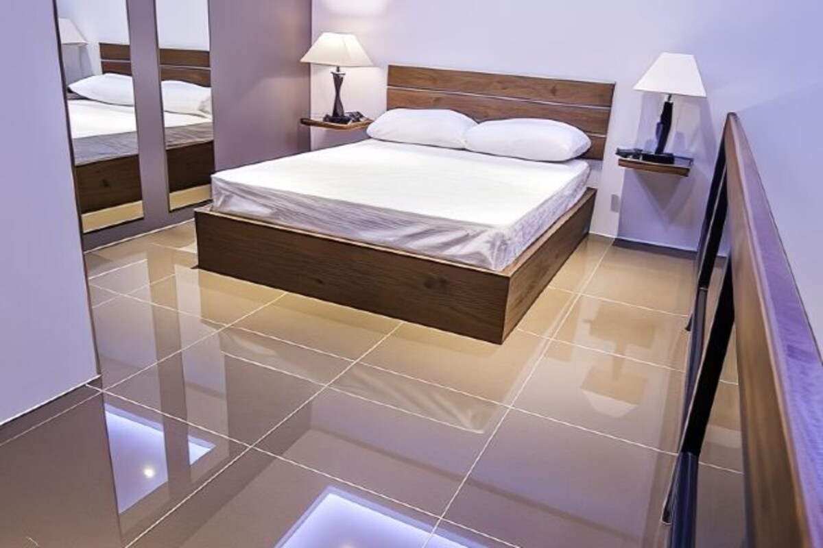 Quarto de hotel moderno com estrutura de cama em madeira, duas mesas de cabeceira com abajures e um espelho grande.