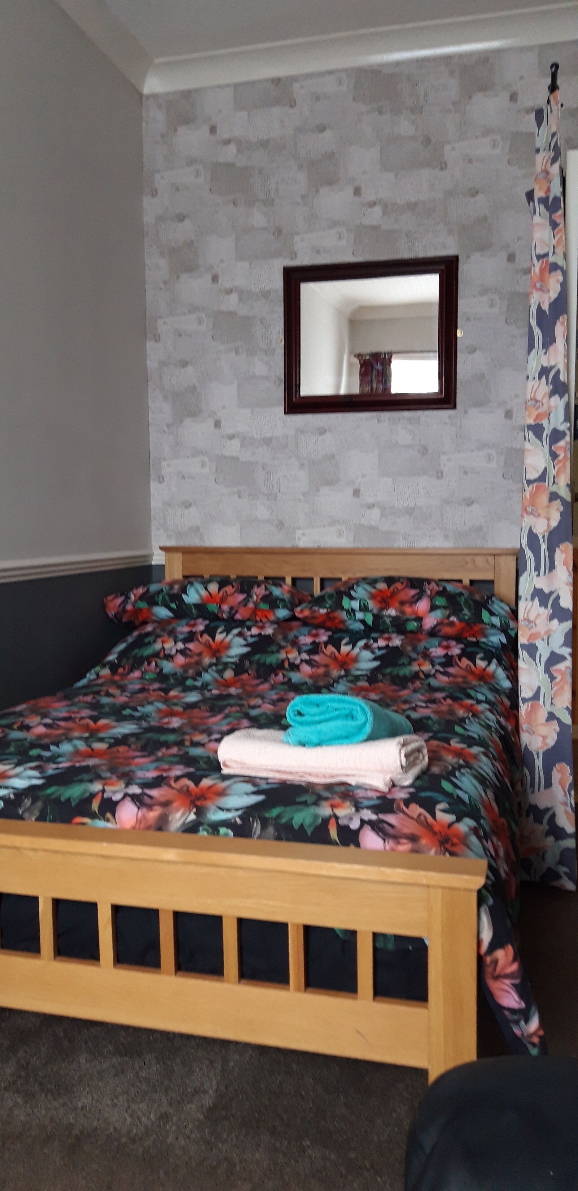 Quarto com cama de madeira, colcha floral, espelho sobre a cama e parede de tijolos cinzas.
