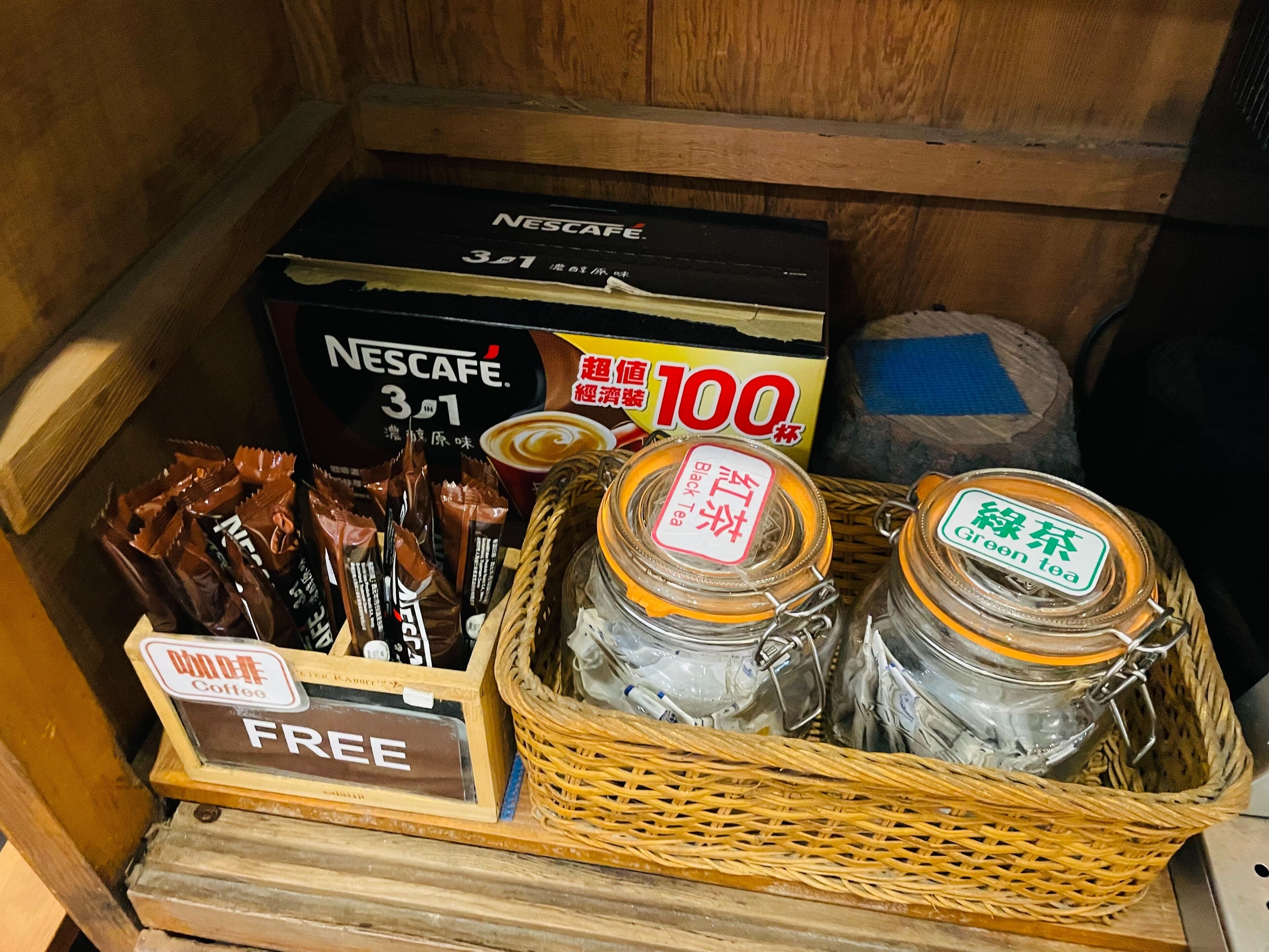 Um espaço de armazenamento com cápsulas de café Nescafé 3 em 1, um aviso indicando café gratuito e dois potes de vidro identificados com tipos de chá.
