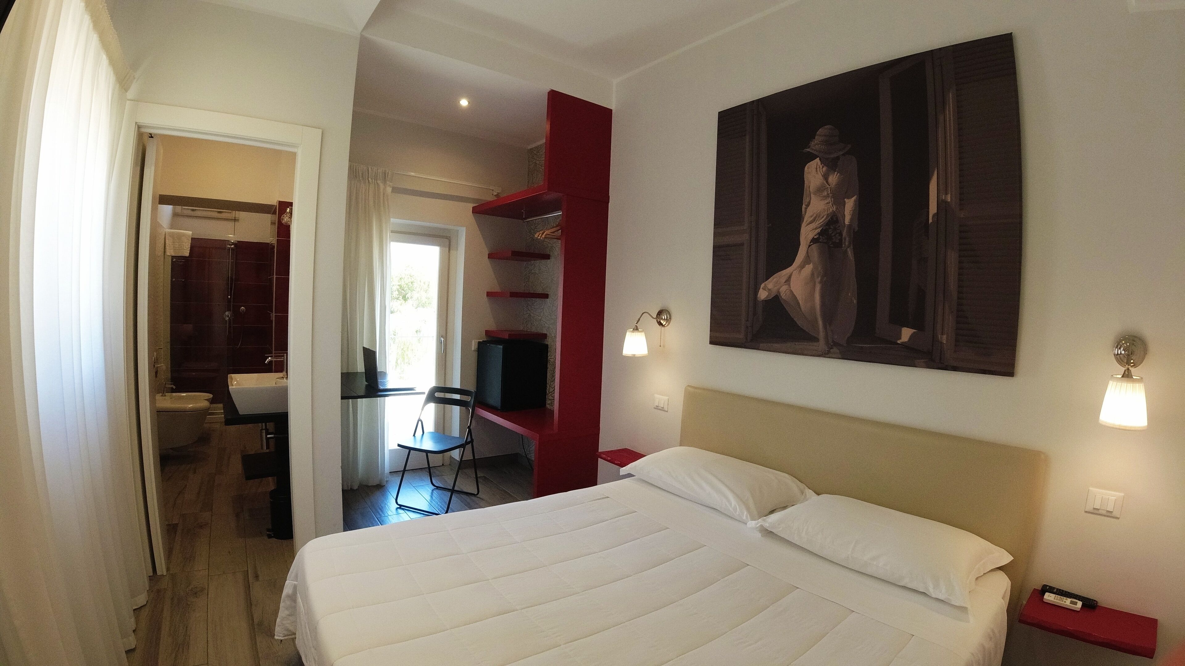 Quarto de hotel com uma cama, uma escrivaninha com um laptop, uma cadeira, área de chuveiro e uma parede com detalhes vermelhos.