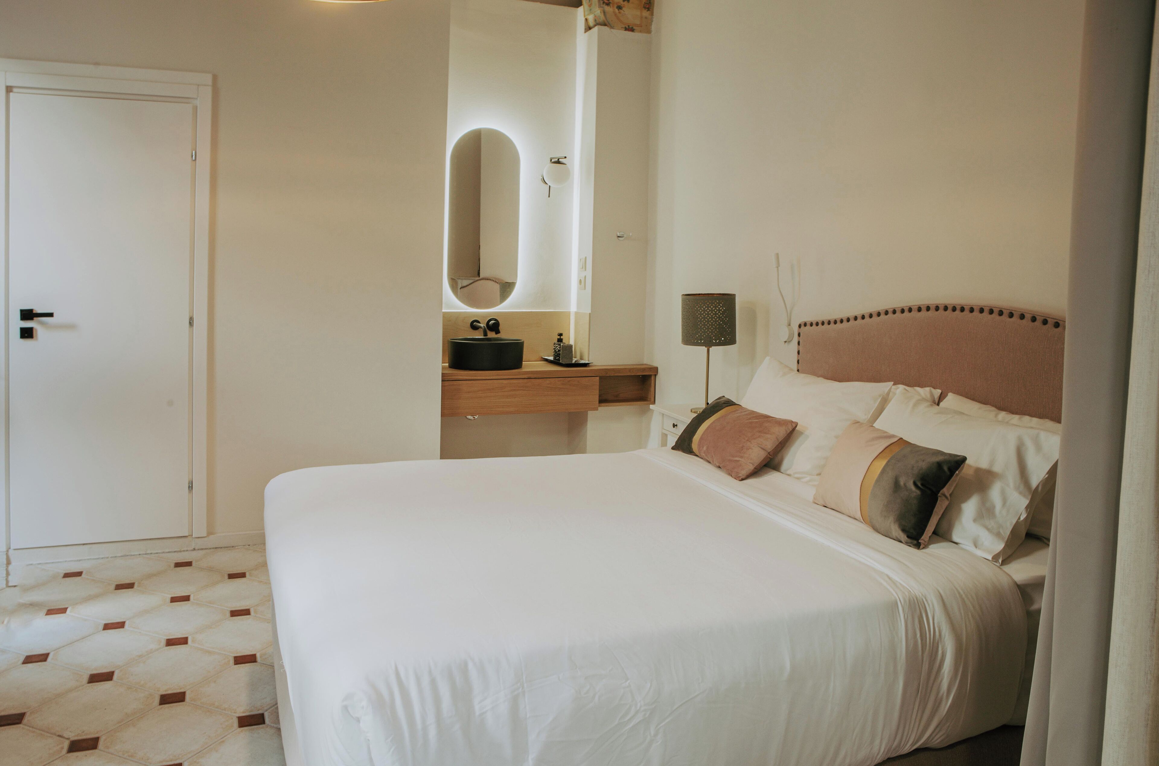 Quarto de hotel com uma cama grande, mesa de cabeceira, espelho, pia e porta.