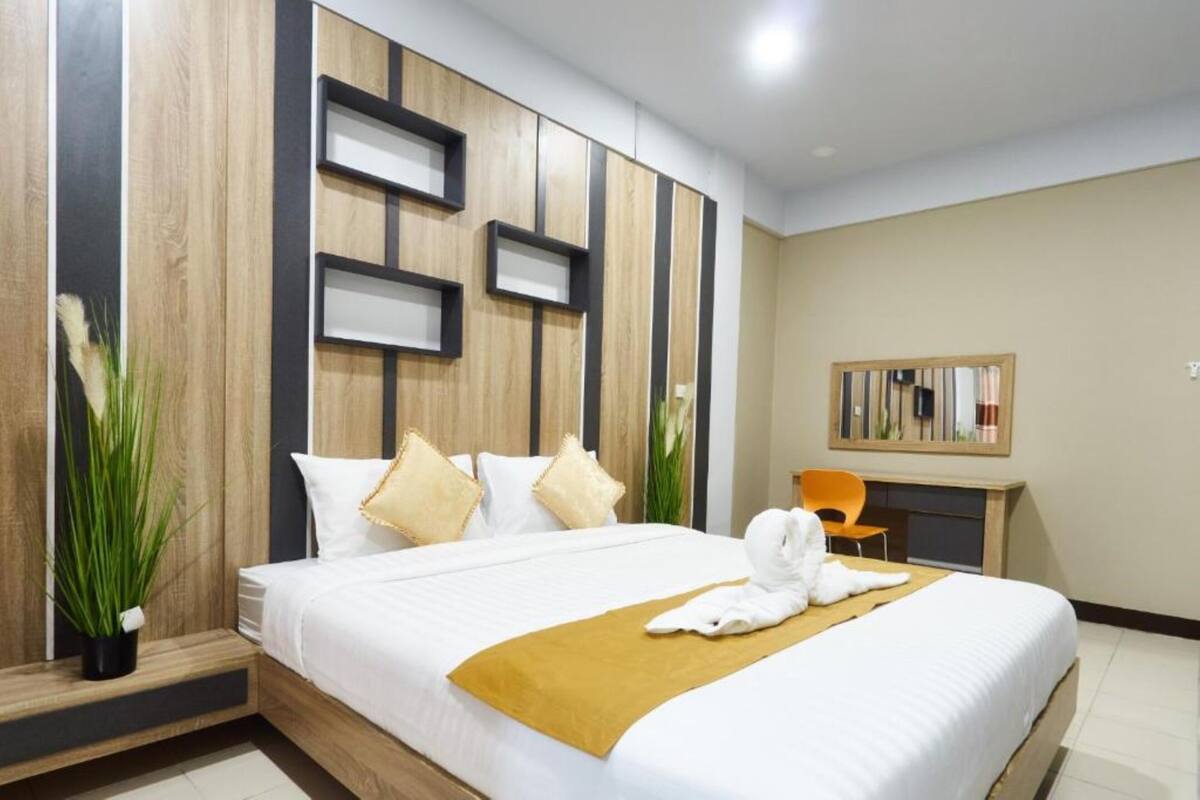 Quarto de hotel com uma cama grande, uma escrivaninha e uma cadeira. Há duas plantas em vasos no chão.