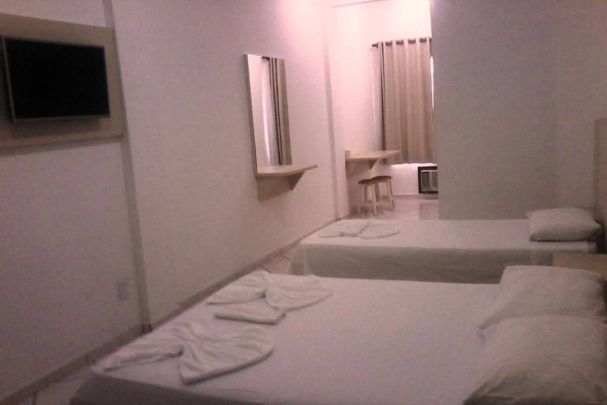 Quarto de hotel com duas camas, televisão fixada na parede, uma mesinha e uma cadeira.
