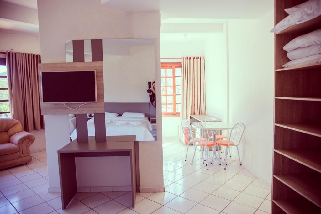 Quarto de hotel com cama, televisão, escrivaninha, cadeira, mesa de jantar com cadeiras e guarda-roupa.