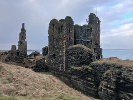 Ruínas de pedra históricas com uma torre desabada, cercadas por terreno gramado e o mar ao fundo.
