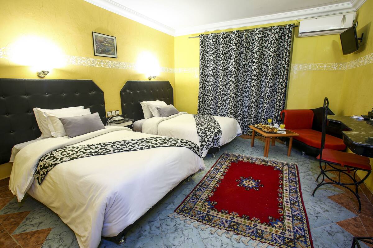 Quarto de hotel com duas camas, um tapete vermelho, uma mesa pequena e uma televisão.
