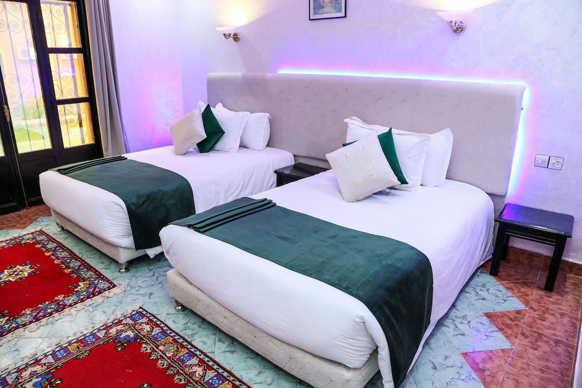 Quarto de hotel com duas camas, um tapete com padrão vermelho e azul, e uma pequena mesa.