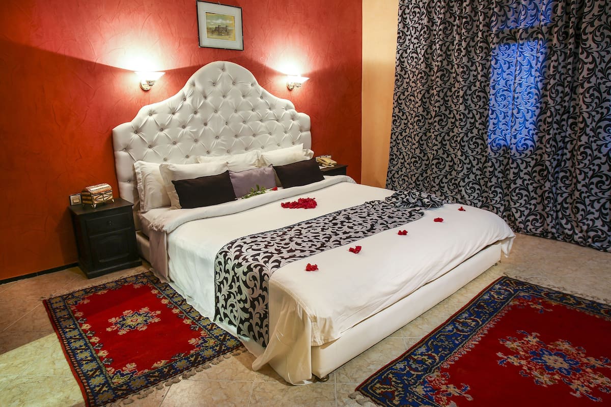 Quarto com cabeceira estofada, colcha de cama estampada em vermelho e preto, e um arranjo floral vermelho sobre a cama.