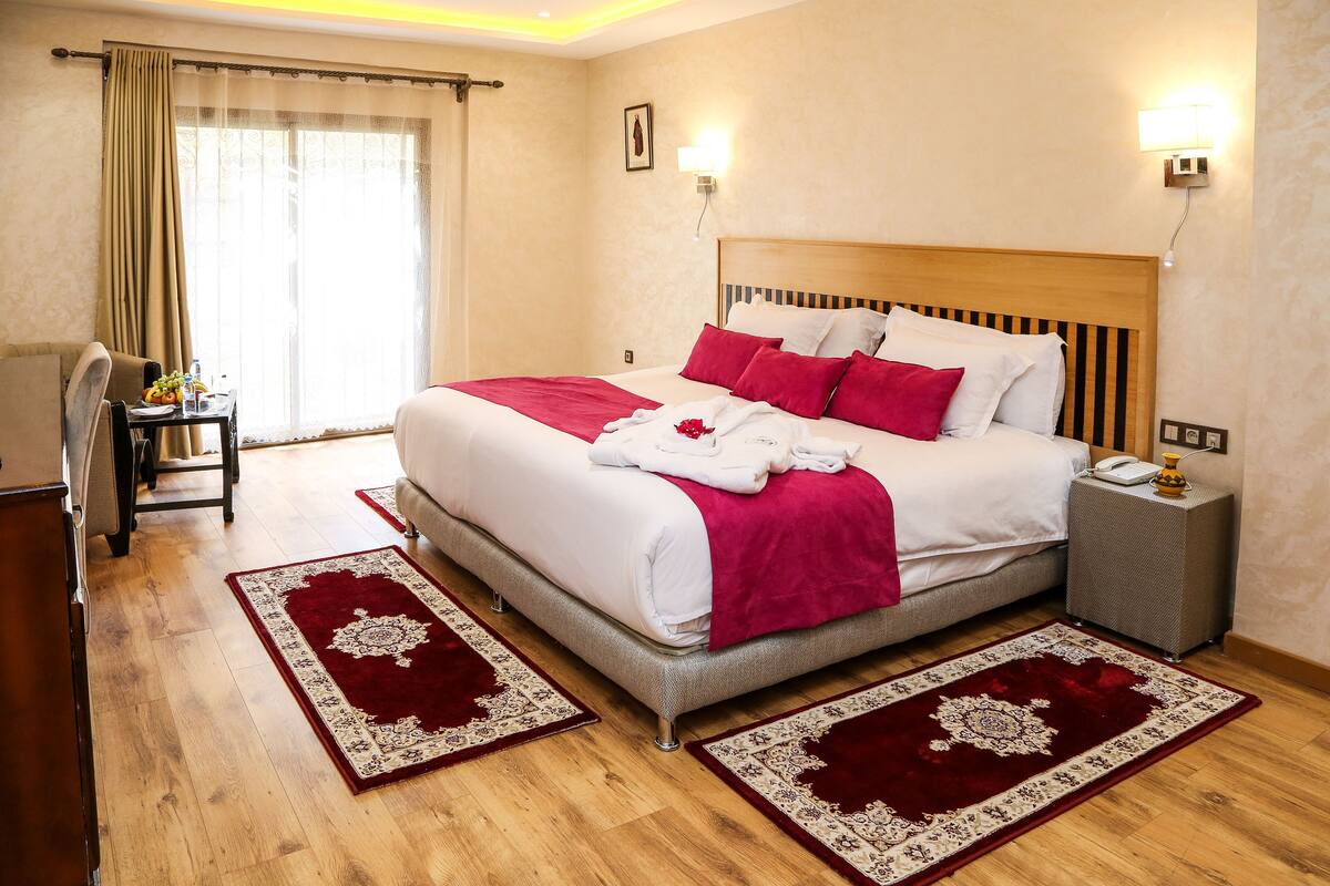 Quarto de hotel com uma cama grande, duas mesas de cabeceira, uma escrivaninha com cadeira, uma janela com cortinas e uma luminária na parede.