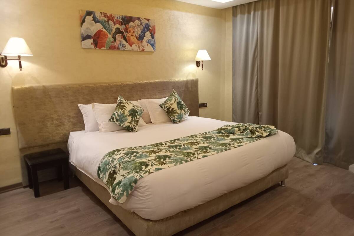 Quarto de hotel com uma cama grande, duas luminárias de cabeceira, um quadro na parede e uma janela com cortinas.
