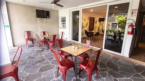 Uma área de refeições com cadeiras vermelhas e uma mesa de madeira, piso de pedra, um ventilador de teto e um extintor de incêndio.