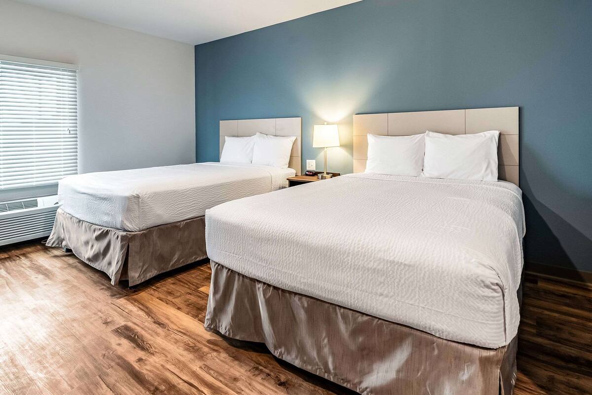 Quarto de hotel com duas camas, uma mesa de cabeceira com abajur, janela com persianas e piso de madeira.