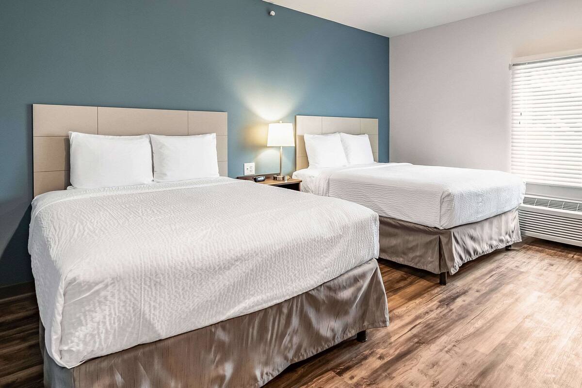 Quarto de hotel com duas camas, uma mesa de cabeceira com luminária e uma janela com persianas.