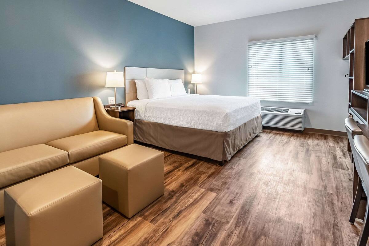Quarto de hotel com cama, sofá, mesa de centro, mesa de cabeceira, abajur, janela com persianas e ar condicionado instalado.