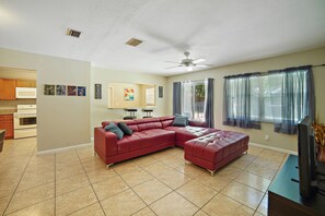Uma sala de estar ampla com um sofá seccional vermelho, um ventilador de teto e uma cozinha ao fundo.