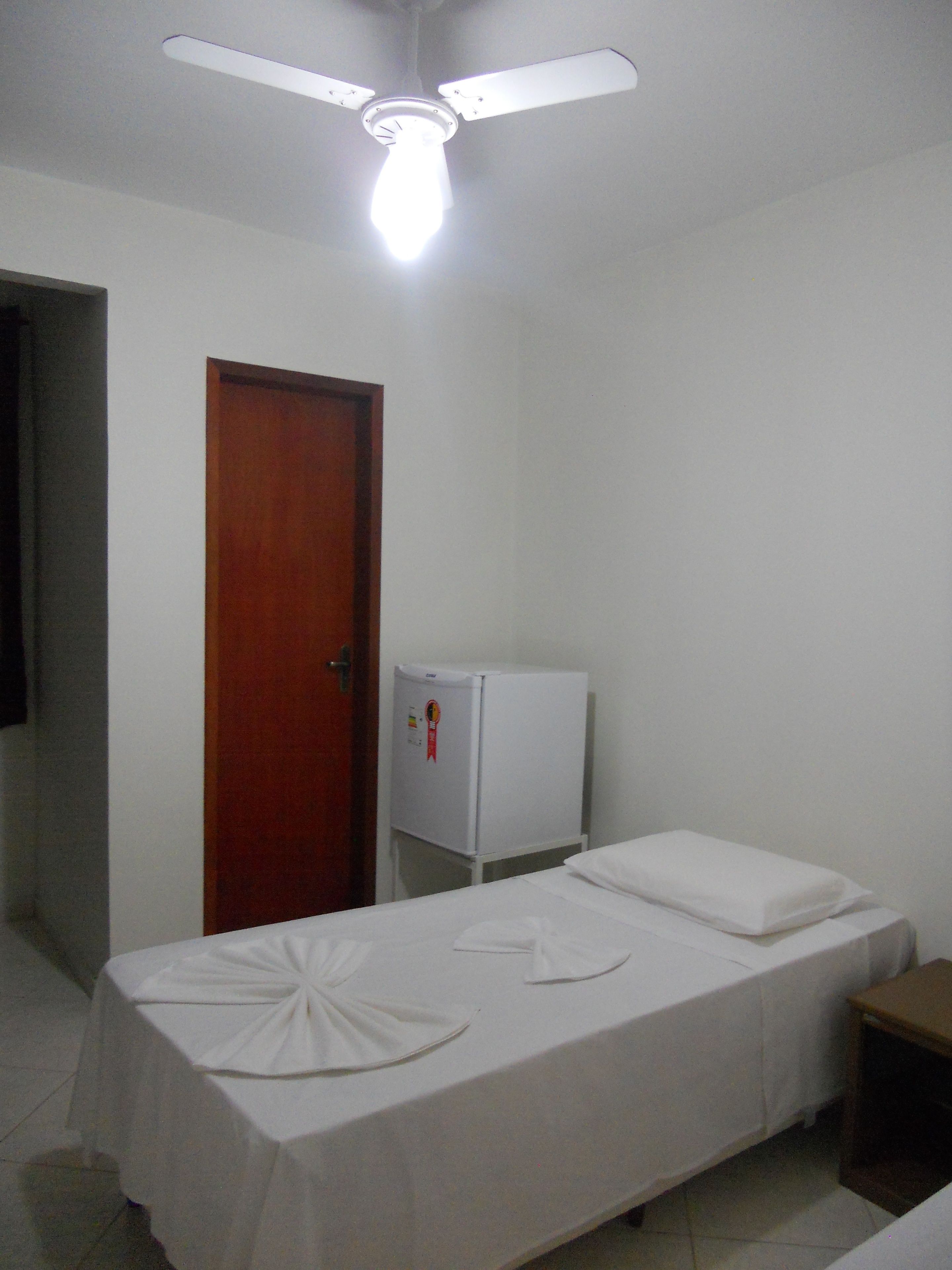 Um quarto pequeno e simples com uma cama de solteiro, uma porta de madeira, um frigorífico branco e um ventilador de teto.