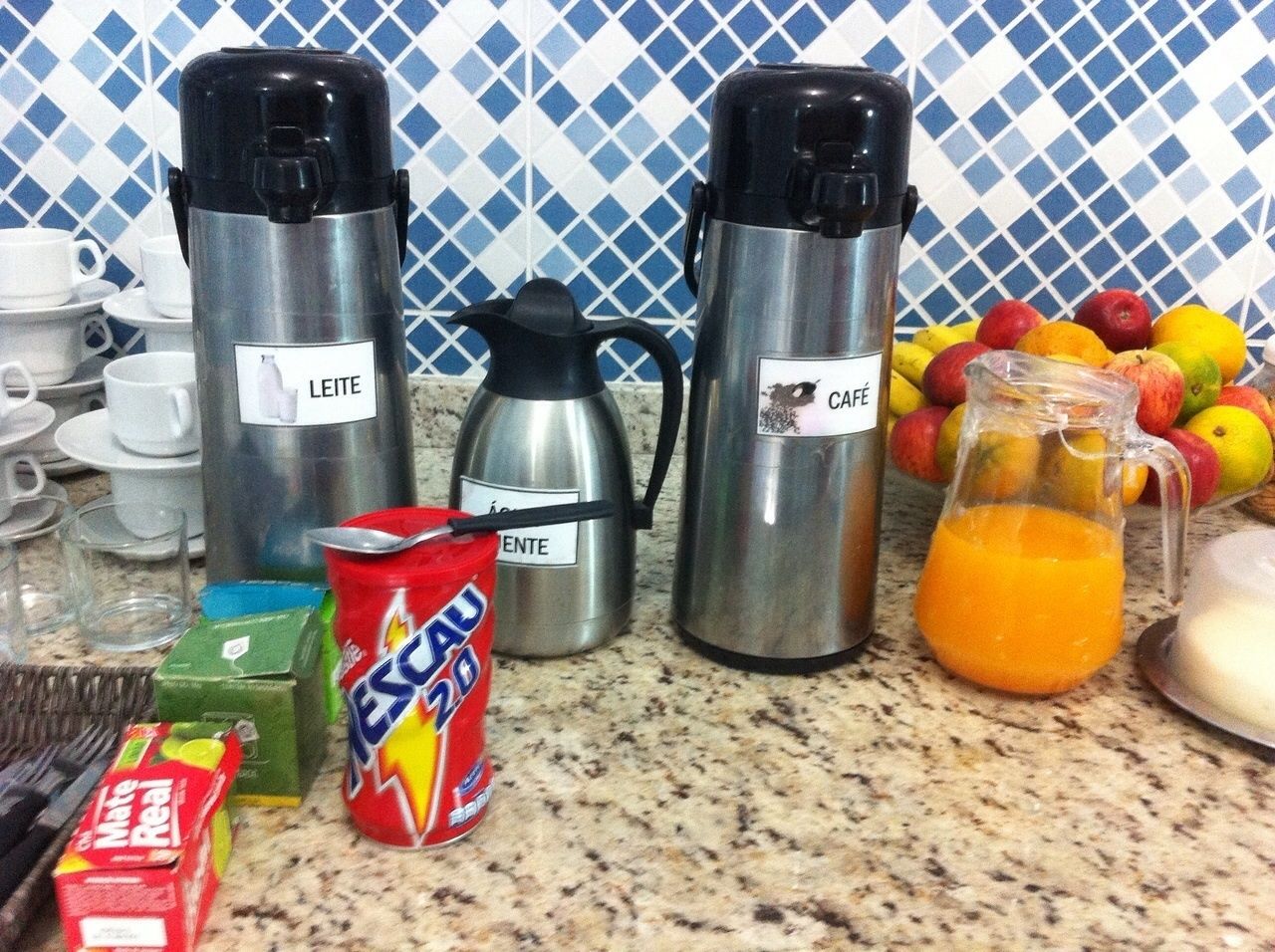 Uma bancada de cozinha com duas garrafas térmicas de aço inoxidável, uma delas identificada com 'LEITE' e a outra com 'CAFÉ'. Há também uma lata vermelha de 'ESCOLA 20', uma caixa verde e um jarro de vidro com suco de laranja.