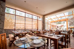 Um restaurante com várias mesas e cadeiras, janelas amplas e uma parede de pedra.