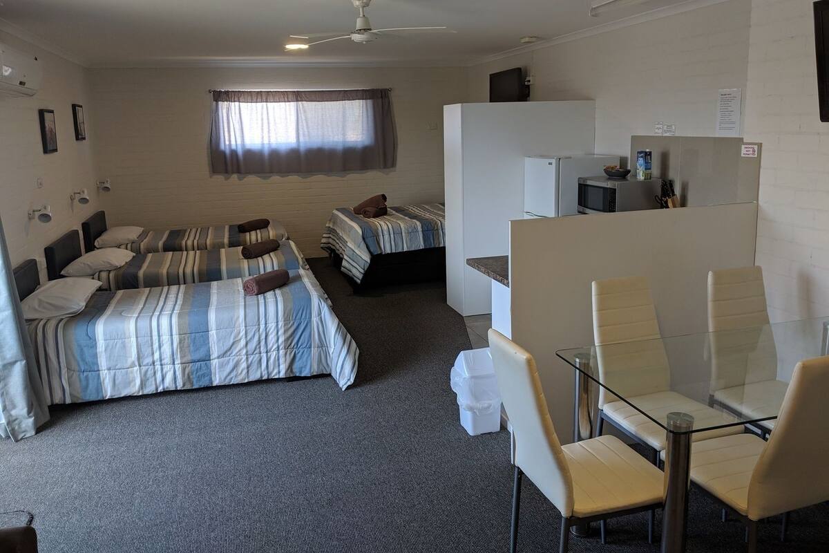 Quarto de hotel com duas camas, área de refeições com mesa de vidro e cozinha compacta.