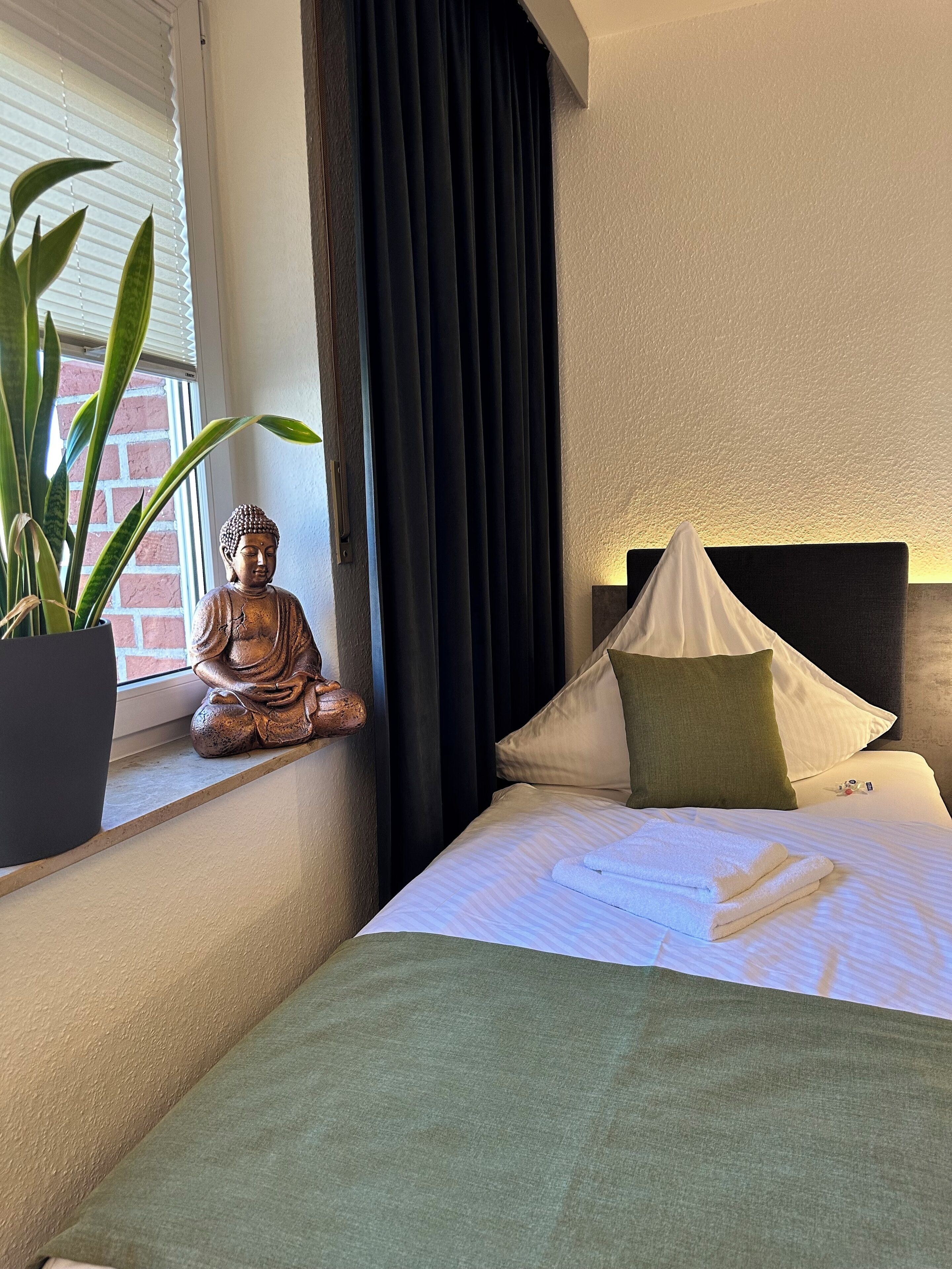 Cama bem arrumada com lençóis brancos e manta verde, uma estátua de Buda em um parapeito e uma planta em vaso ao lado da janela.