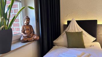 Cama bem arrumada com lençóis brancos e manta verde, uma estátua de Buda em um parapeito e uma planta em vaso ao lado da janela.