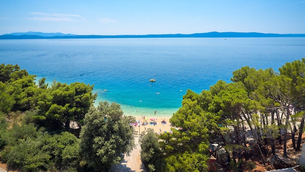Uma praia com águas azul-claras, rodeada por árvores verdes e montanhas ao fundo.