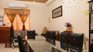 Uma área de jantar com mesa de vidro e cadeiras pretas, um sofá com almofadas estampadas e uma parede decorativa com luminárias pendentes.