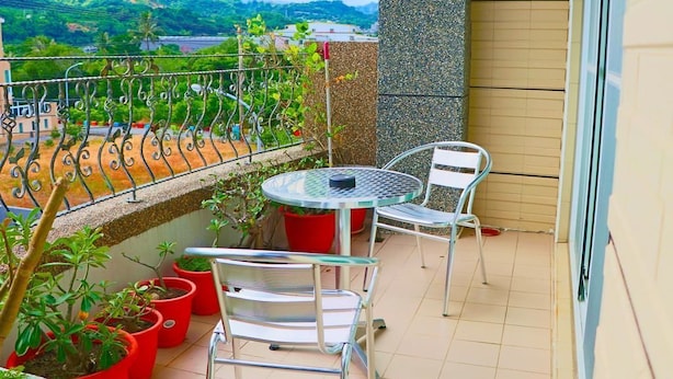 Varanda com mesa redonda e duas cadeiras, plantas em vasos e vista para colinas verdes.