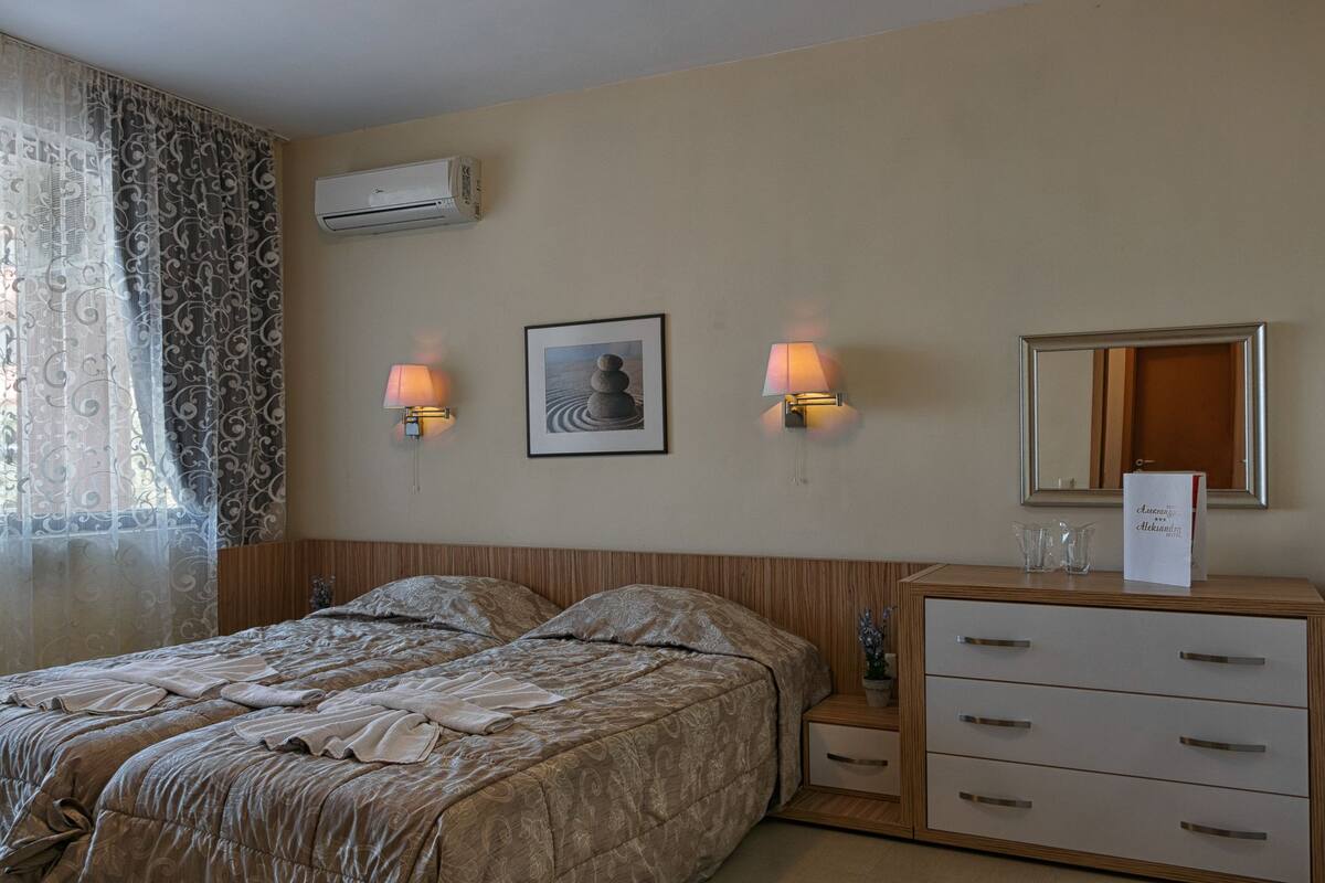 Quarto de hotel com duas camas, uma mesa de cabeceira com abajur, ar-condicionado fixado na parede, um quadro emoldurado e um espelho.