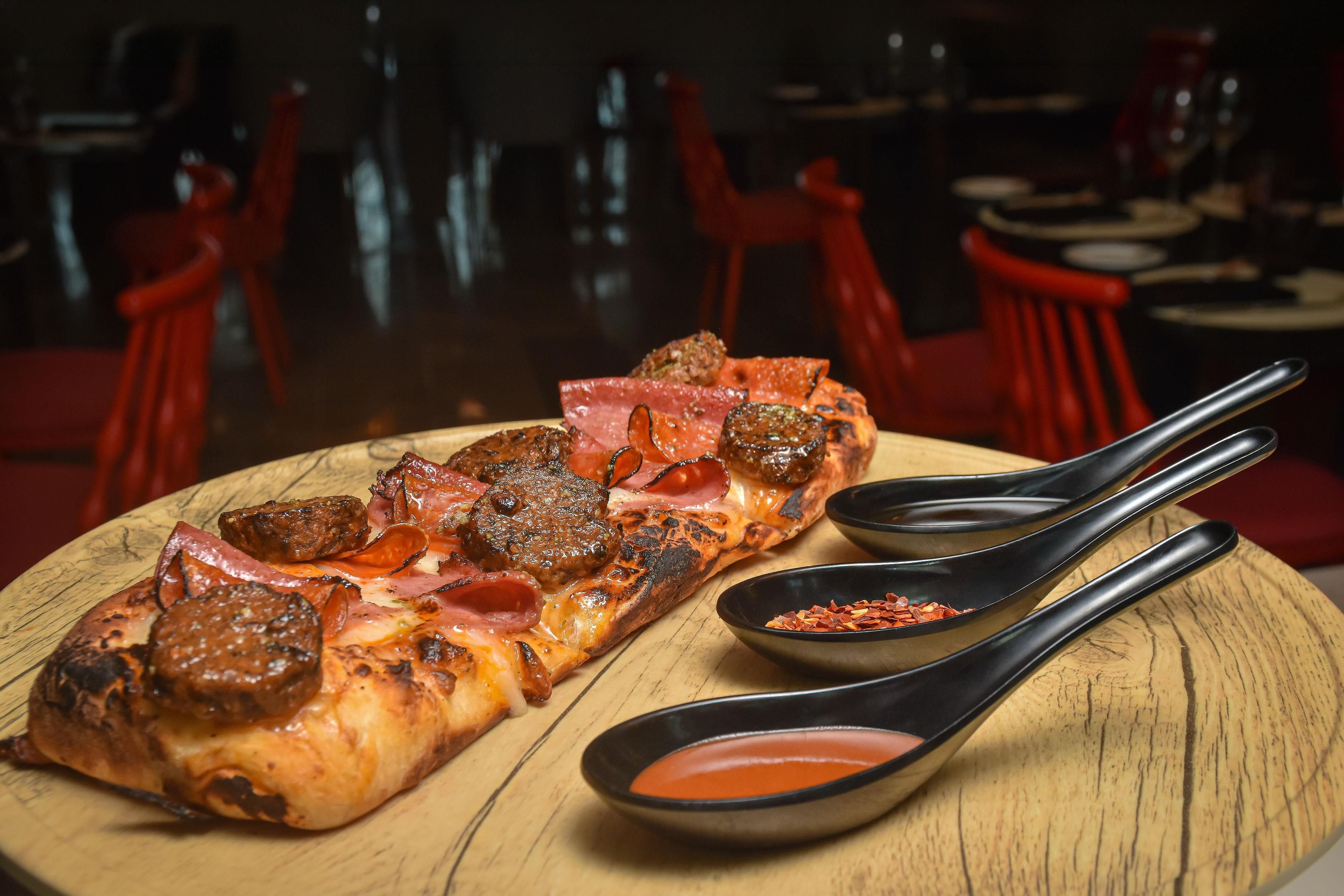 Pizza com diversos ingredientes sobre uma mesa de madeira, acompanhada por duas colheres pretas e uma tigela de molho.