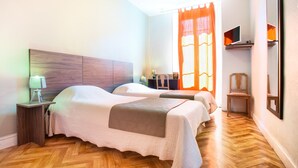 Quarto de hotel com duas camas, piso de madeira, mesa de cabeceira com abajur, uma cadeira e televisão na parede.