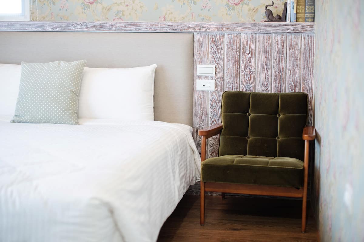 Cama bem arrumada com lençóis brancos e uma poltrona estofada verde com braços de madeira.