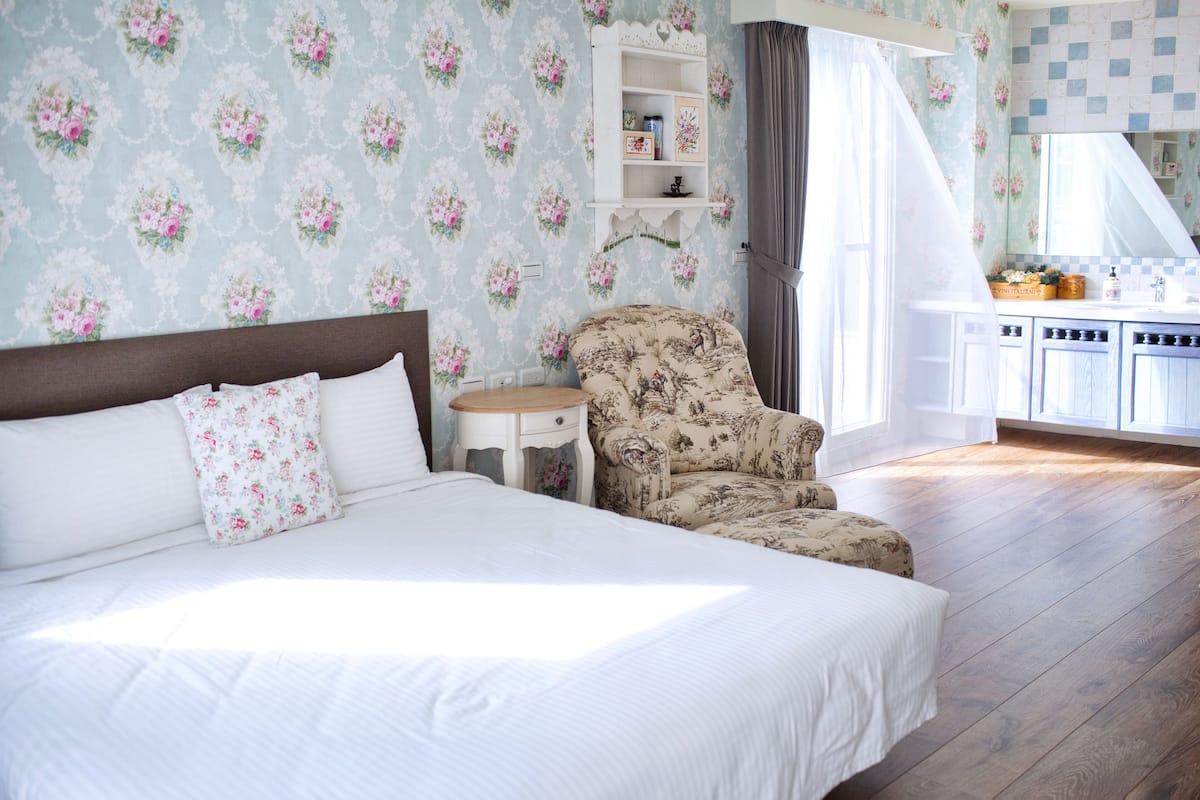 Um quarto com uma cama grande, uma poltrona com padrão floral, uma mesinha de cabeceira e uma prateleira branca com objetos decorativos.