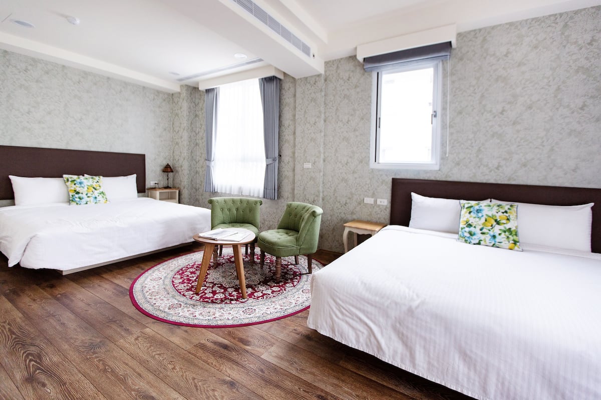 Quarto de hotel com duas camas, uma poltrona verde, uma mesa redonda pequena e um tapete com padrão vermelho e branco.