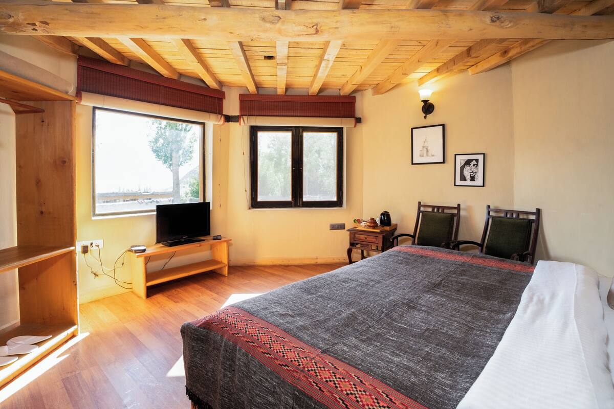 Quarto com cama grande, teto de madeira, televisão em suporte de madeira, janela e escrivaninha com cadeiras.