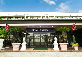 A entrada do Hotel Kazusa, com placa visível, plantas em vasos e uma estátua.