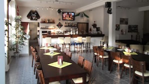 Uma sala de jantar ampla com várias mesas e cadeiras, um espaço bar com balcão e banquetas, e uma cozinha ao fundo.