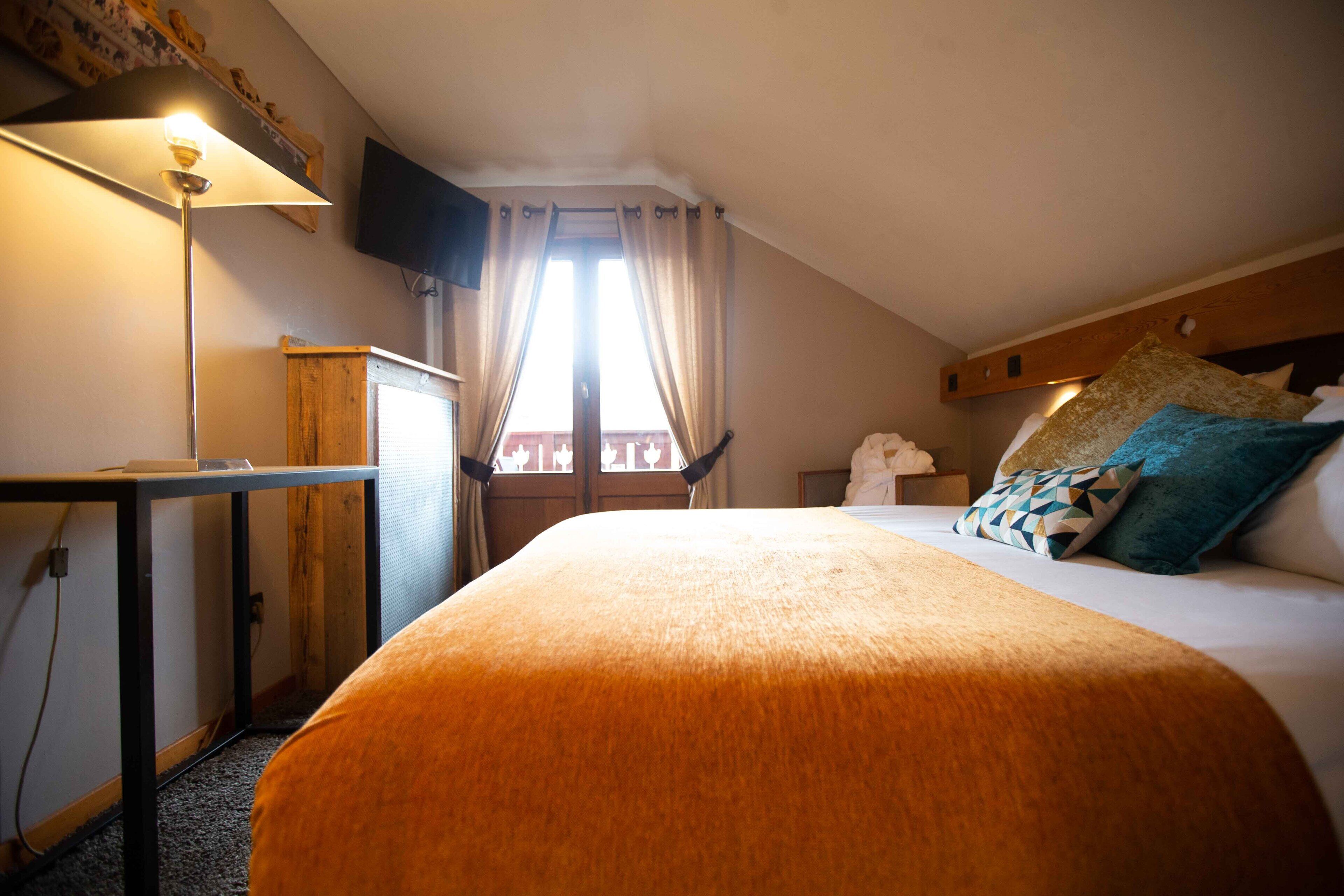 Quarto de hotel com cama, escrivaninha, abajur e televisão na parede.
